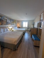 Quarto de hotel com duas camas, uma escrivaninha, uma cadeira, uma televisão e uma janela com cortinas.