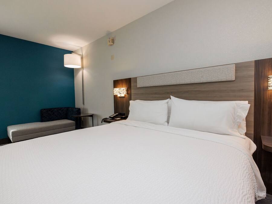 Quarto de hotel com uma cama grande, um banquinho, uma mesinha de cabeceira e uma luminária fixada na parede.