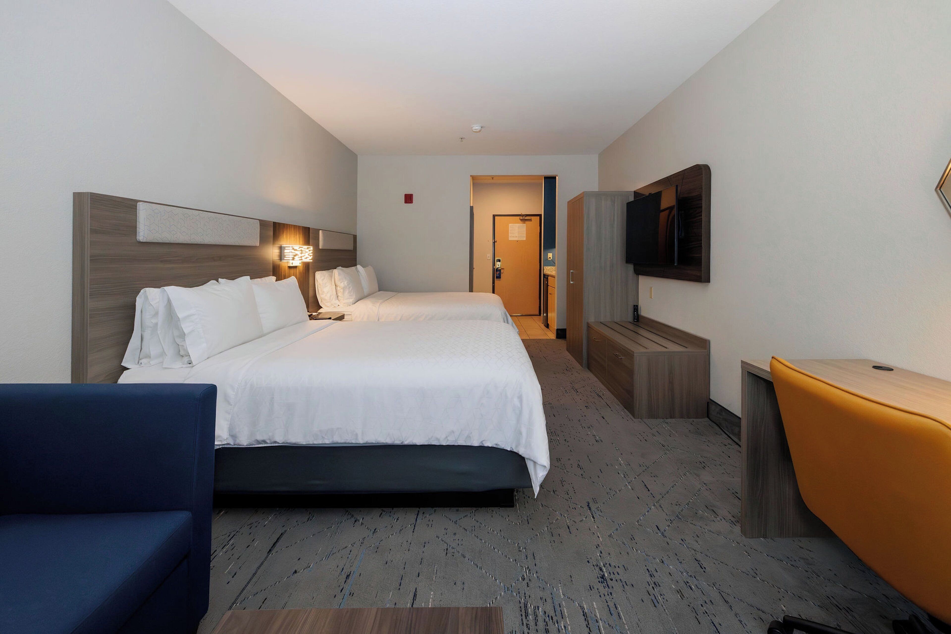 Quarto de hotel com duas camas, uma escrivaninha, uma cadeira, televisão e porta que dá para outro espaço.