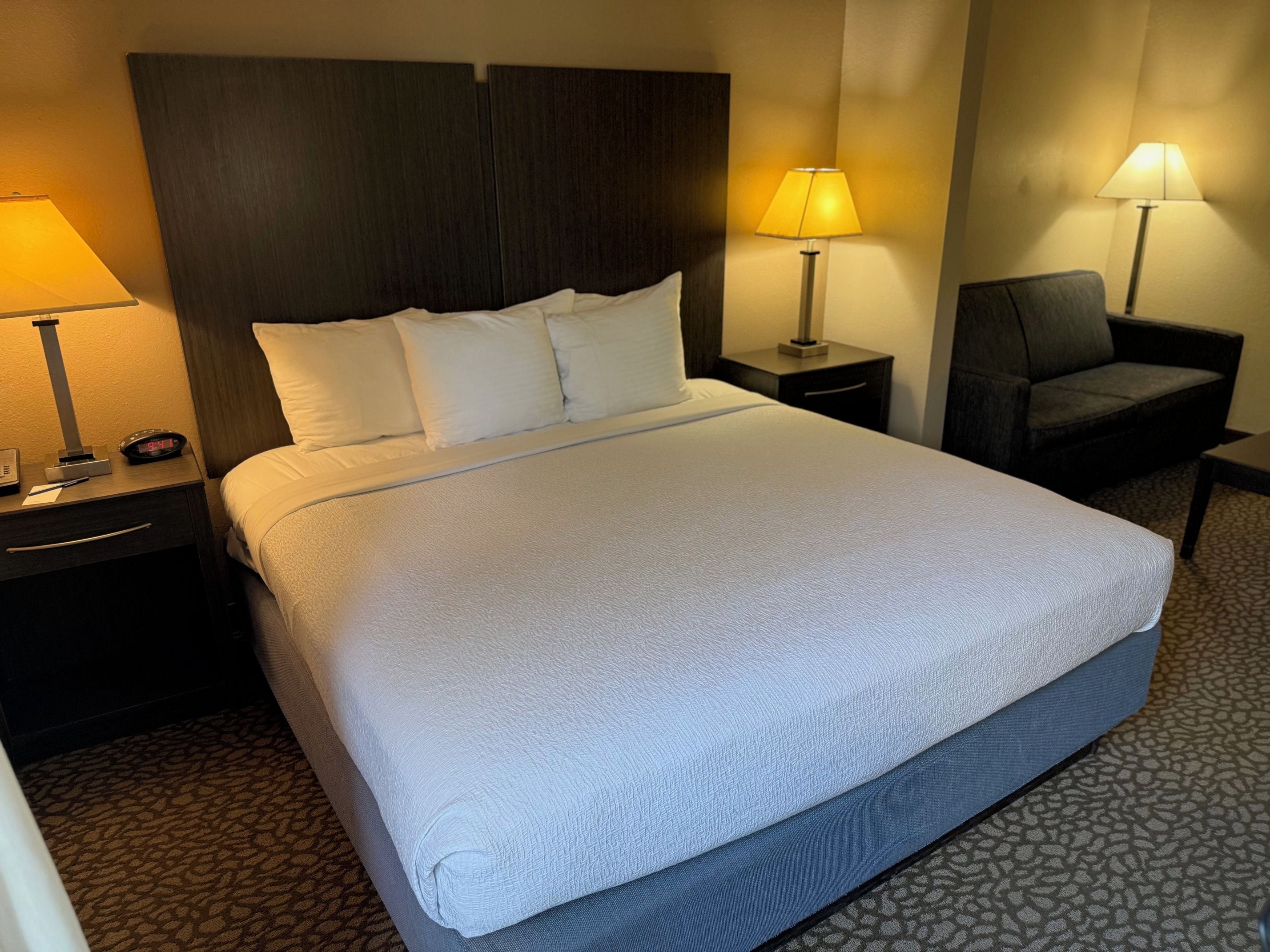 Quarto de hotel com uma cama grande, duas mesas de cabeceira, uma cadeira e duas luminárias.