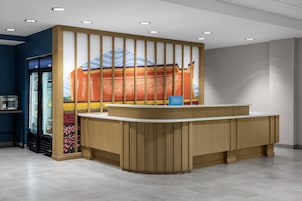 Bancada de recepção com acabamento em madeira, divisória de vidro e placa azul com o nome do hotel.