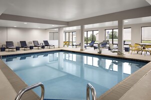 Piscina com espreguiçadeiras e mesas, rodeada por grandes janelas.