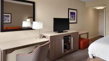Quarto de hotel com cama, escrivaninha com televisão, cadeira, espelho e porta.