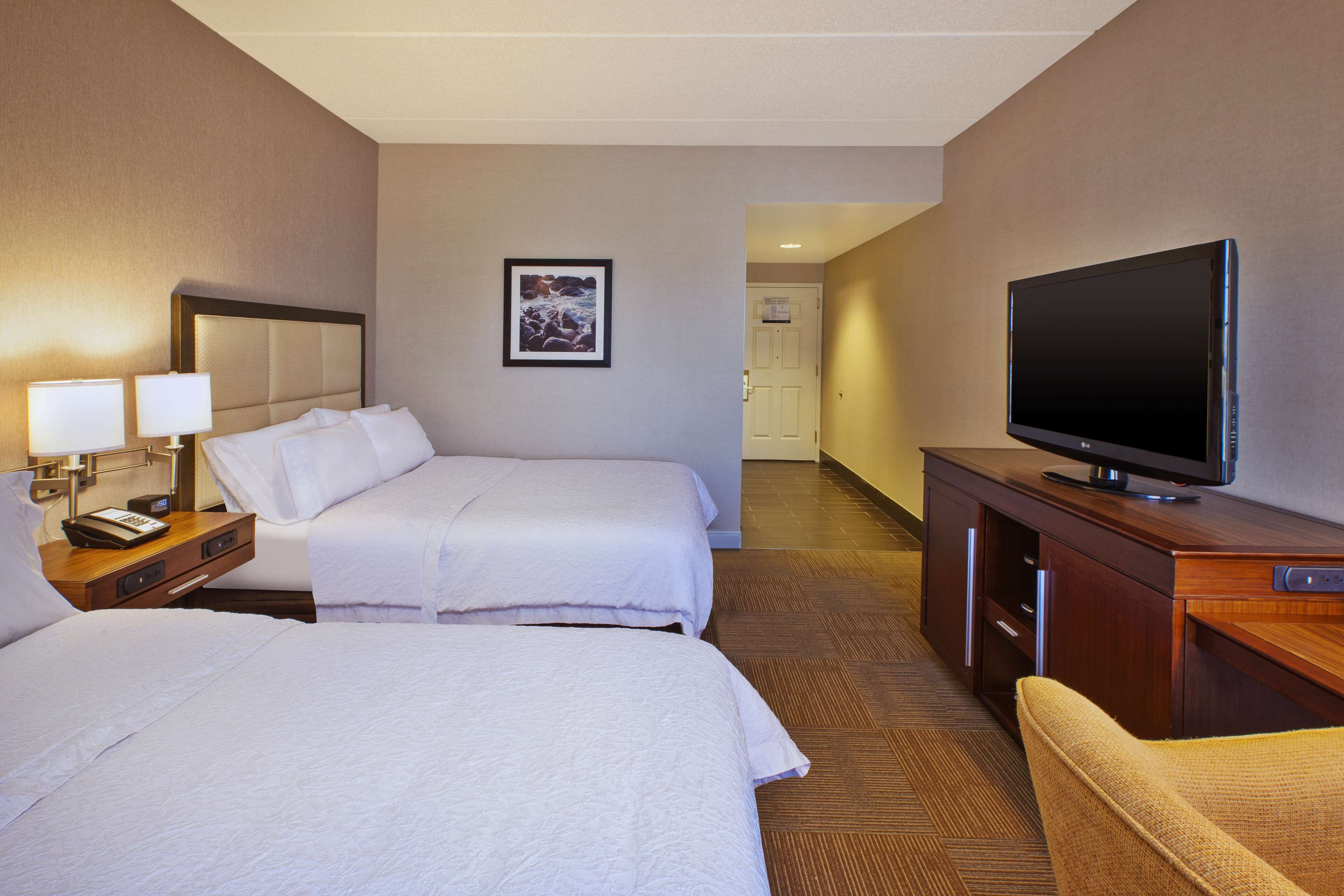 Quarto de hotel com duas camas, televisão, escrivaninha com luminária e um quadro na parede.