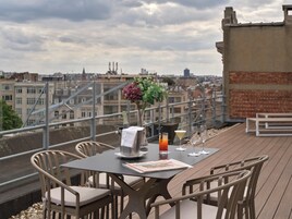 Terraço no telhado com mesa posta para dois, um vaso com flores e um jornal.
