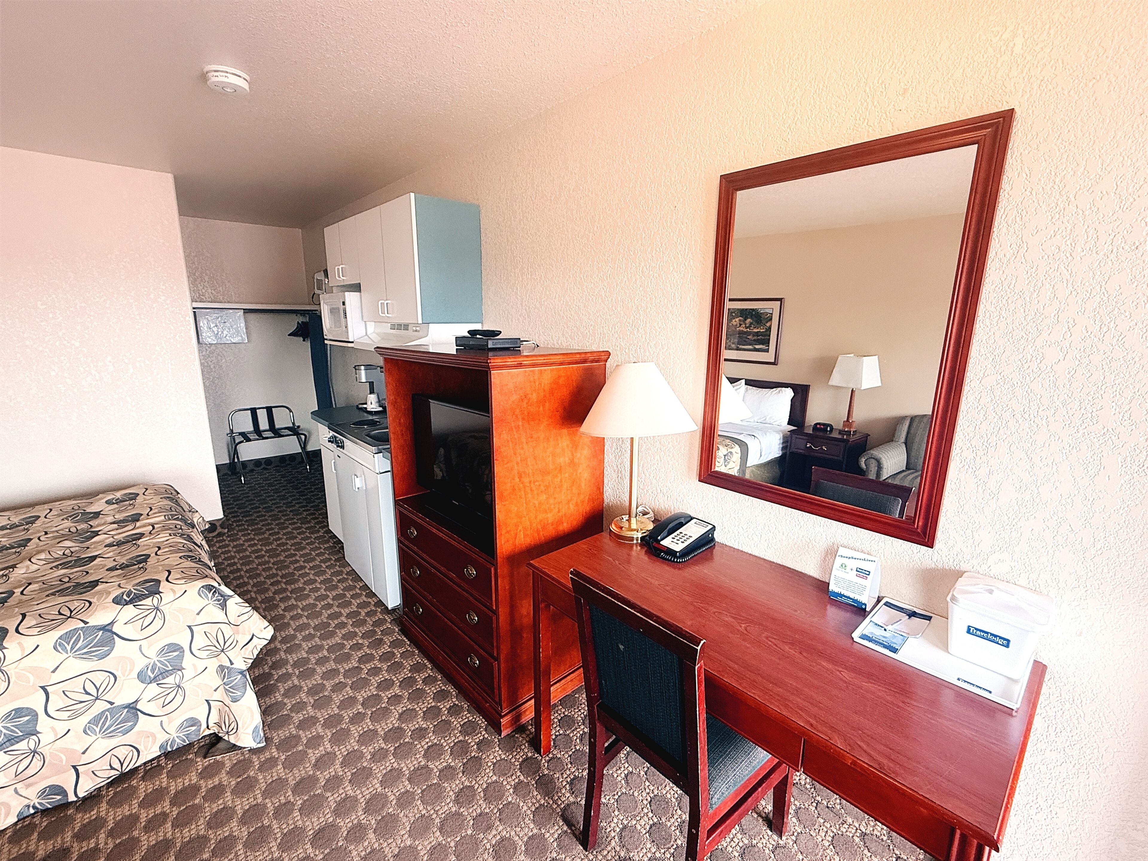 Quarto de hotel com uma cama, escrivaninha, cadeira, espelho, telefone, abajur, micro-ondas e uma pequena cozinha equipada.