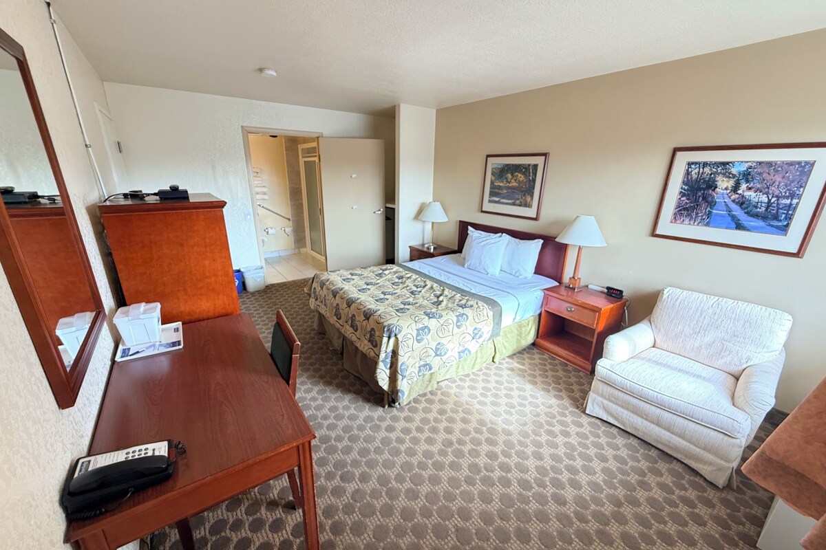 Quarto de hotel com cama, escrivaninha, cadeira, mesa de cabeceira e porta que dá para um corredor.
