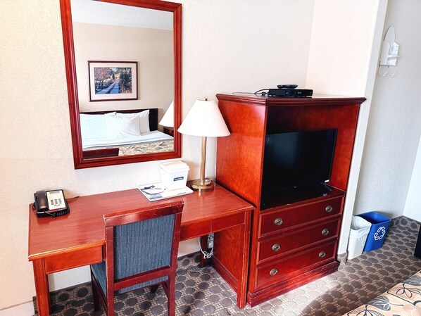 Quarto de hotel com escrivaninha de madeira, uma cadeira, televisão, luminária, cama com roupa branca, quadro na parede e lixeira.