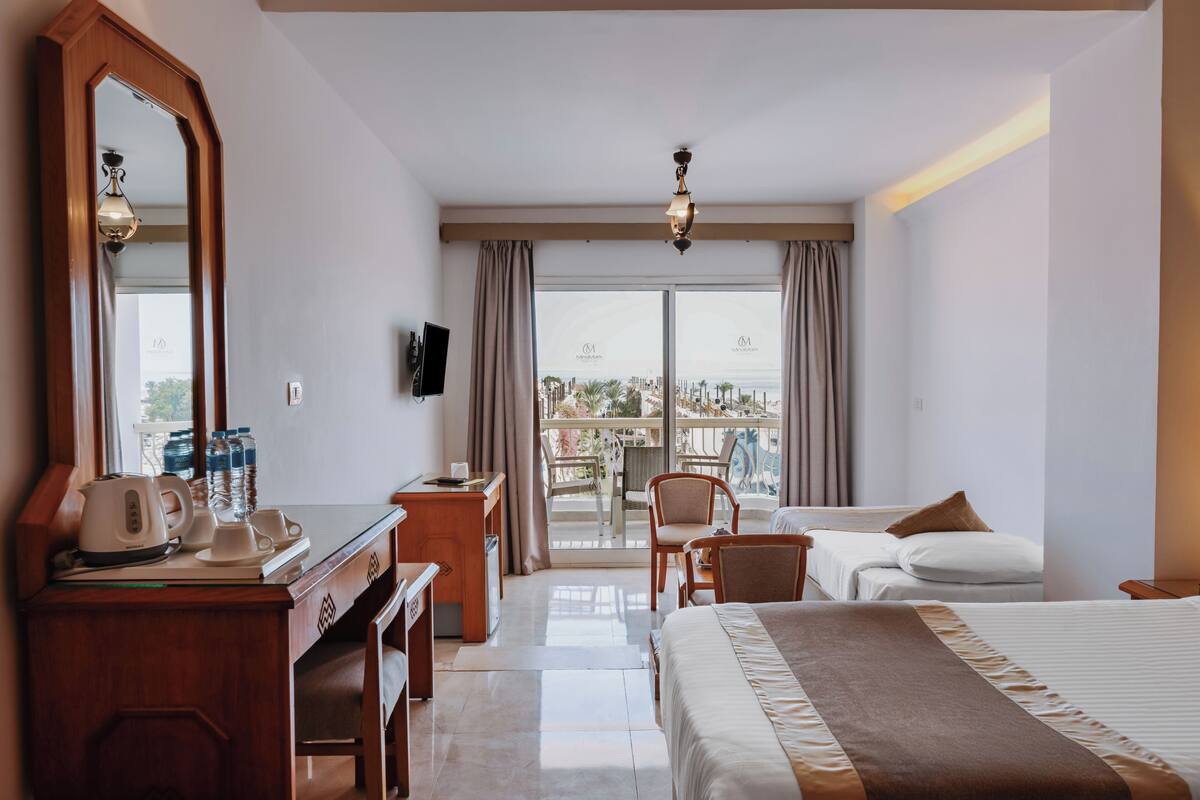 Quarto de hotel com cama, escrivaninha com chaleira e chávenas, uma cadeira, televisão e varanda com vista.