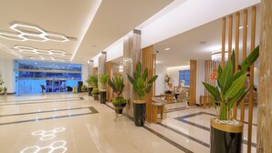 Um lobby moderno com piso lustroso, grandes vasos de plantas e uma área de recepção ao fundo.