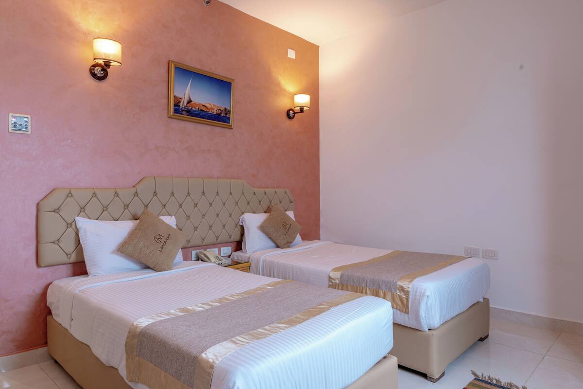 Quarto de hotel com duas camas, cabeceira, luminárias fixadas na parede, um quadro e um telefone.