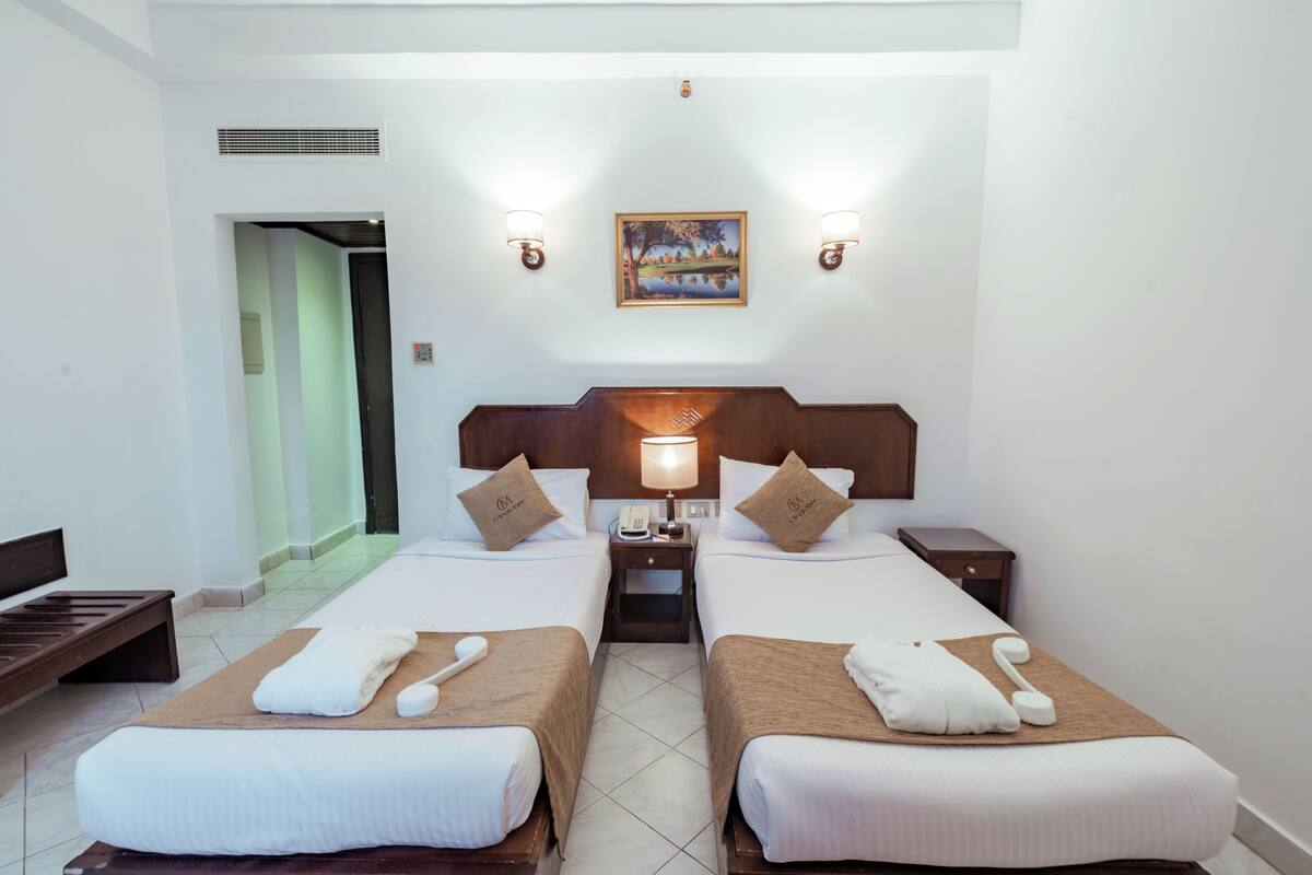 Quarto de hotel com duas camas, uma mesa de cabeceira com abajur, um quadro na parede e uma porta que dá para outro cômodo.