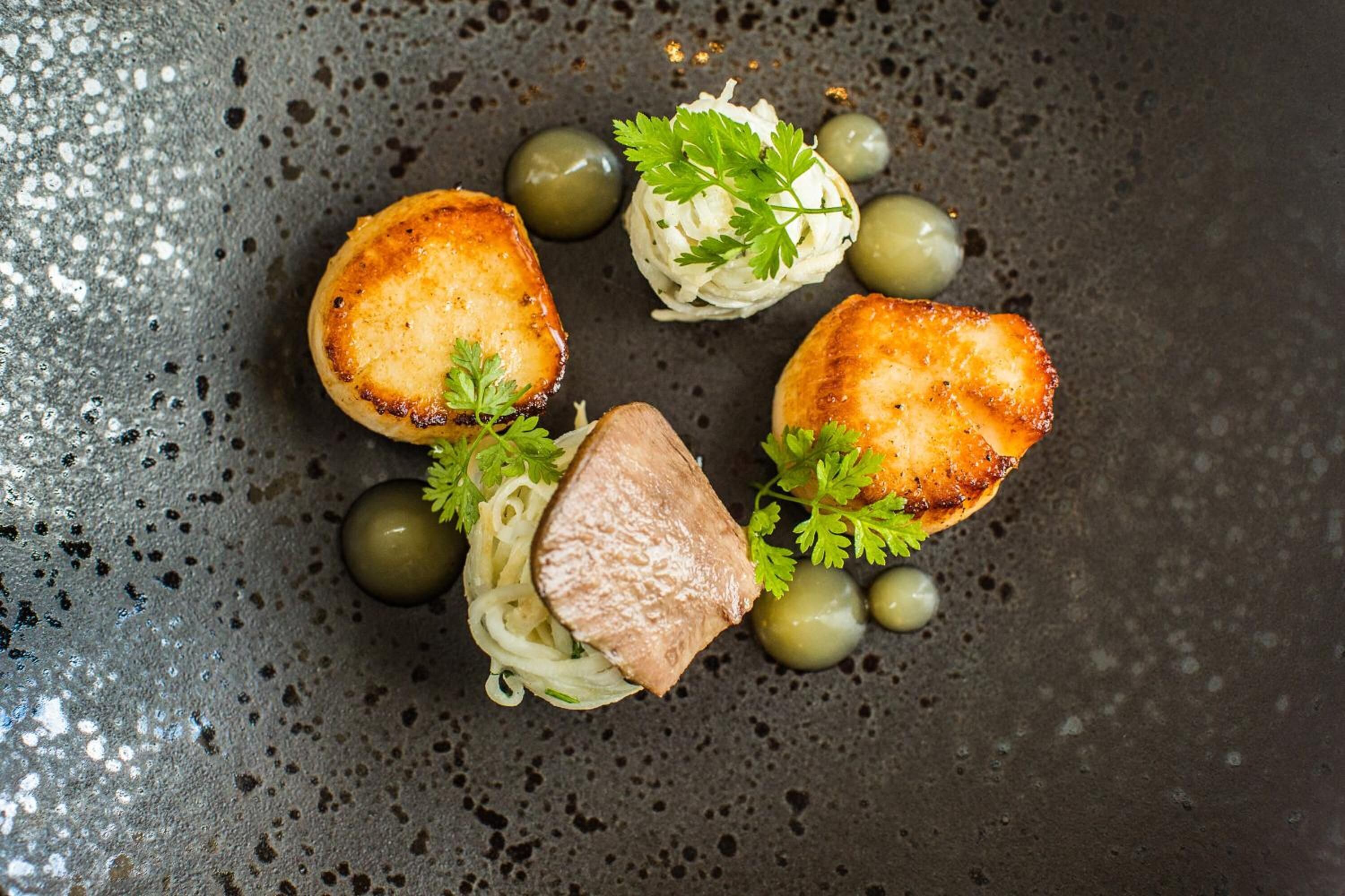 Prato com vieiras grelhadas, um pedaço de carne e enfeite de salsa, servido em prato texturizado com azeite e azeitonas verdes.