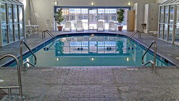 Piscina interior com paredes de vidro transparente, chão de tijolos e várias espreguiçadeiras e plantas em vasos ao redor da área da piscina.