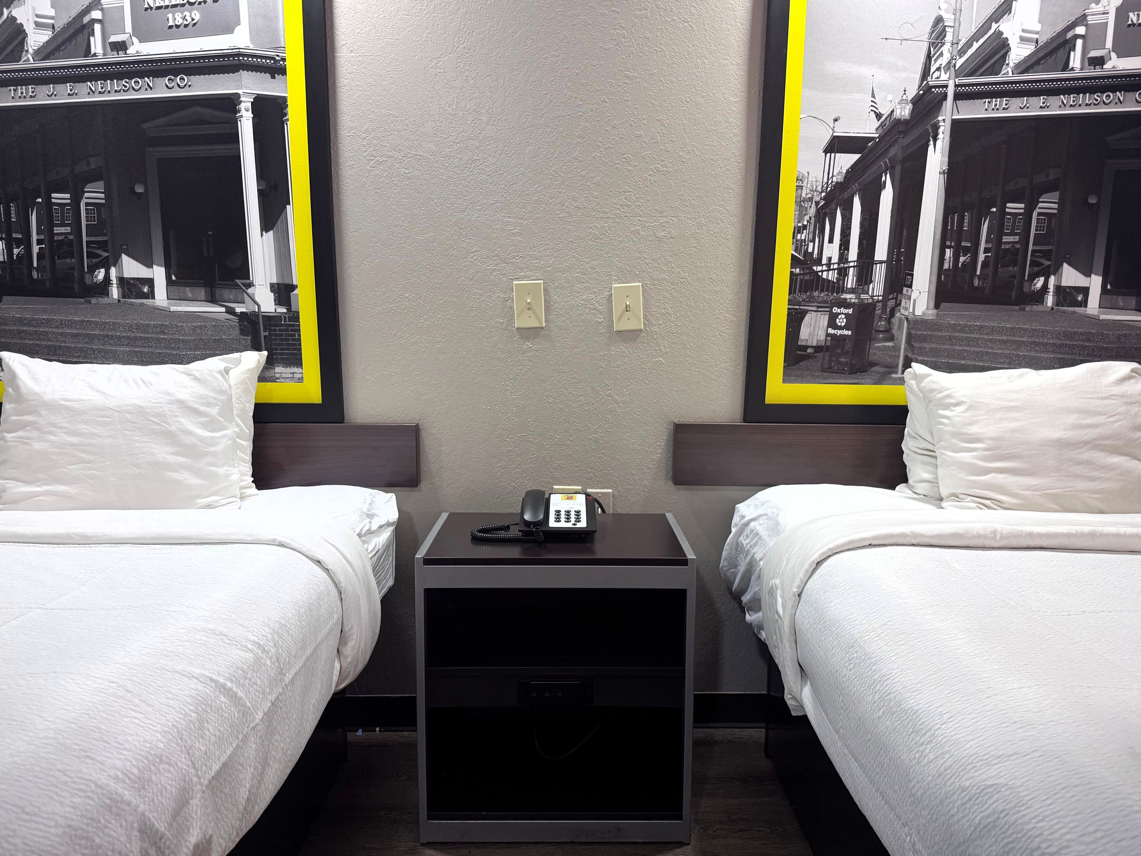 Quarto de hotel com duas camas, uma pequena mesa de cabeceira com telefone e um espelho refletindo o quarto.