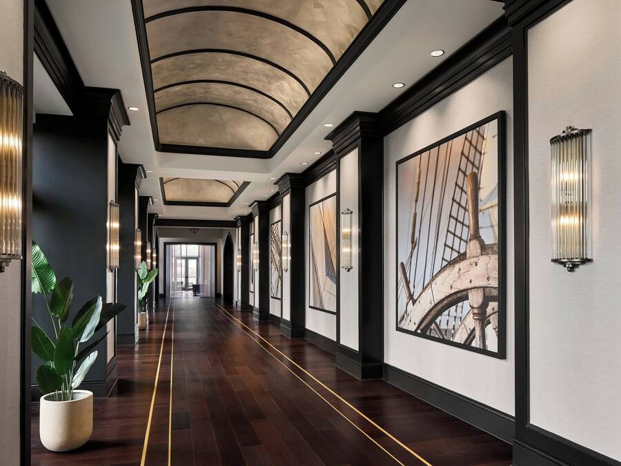 Um corredor comprido e estreito com piso de madeira escura, iluminação embutida e grandes obras de arte emolduradas nas paredes.