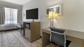 Quarto de hotel com escrivaninha, cadeira, televisão, aparelho de ar condicionado e janela com cortinas.