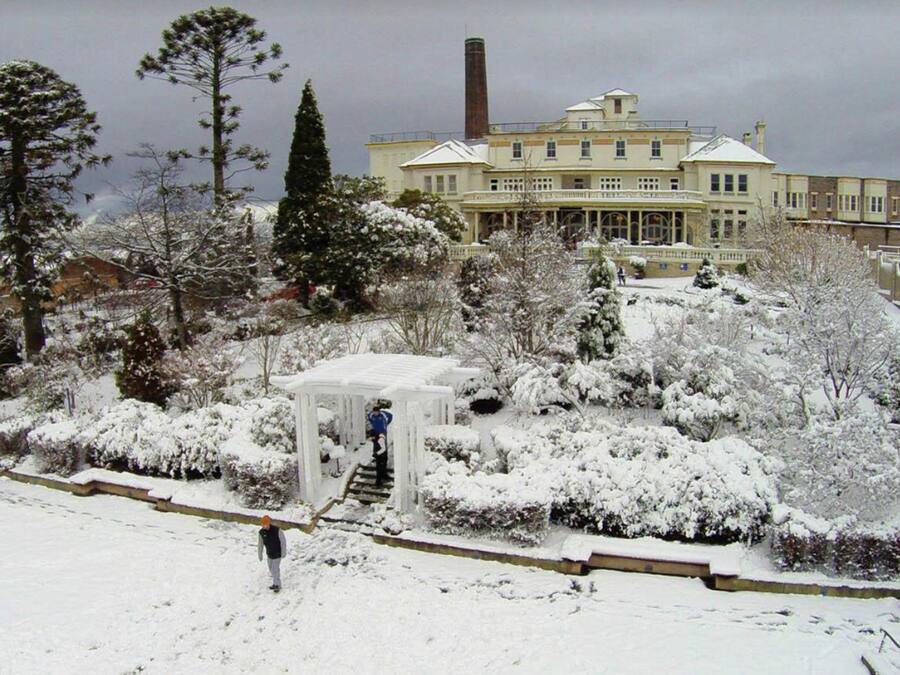 Um edifício grande com uma chaminé notável, rodeado por árvores cobertas de neve e um coreto.