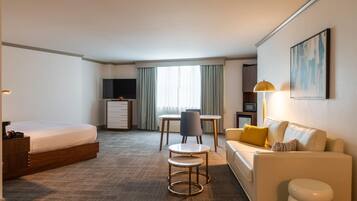 Quarto de hotel com cama, sofá, mesa de jantar com cadeiras, televisão e luminária.