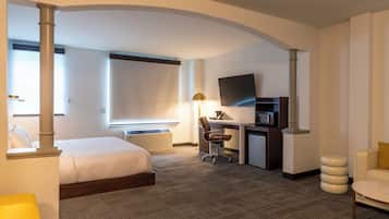 Quarto de hotel com cama, escrivaninha, cadeira, televisão e um pequeno espaço para sentar.