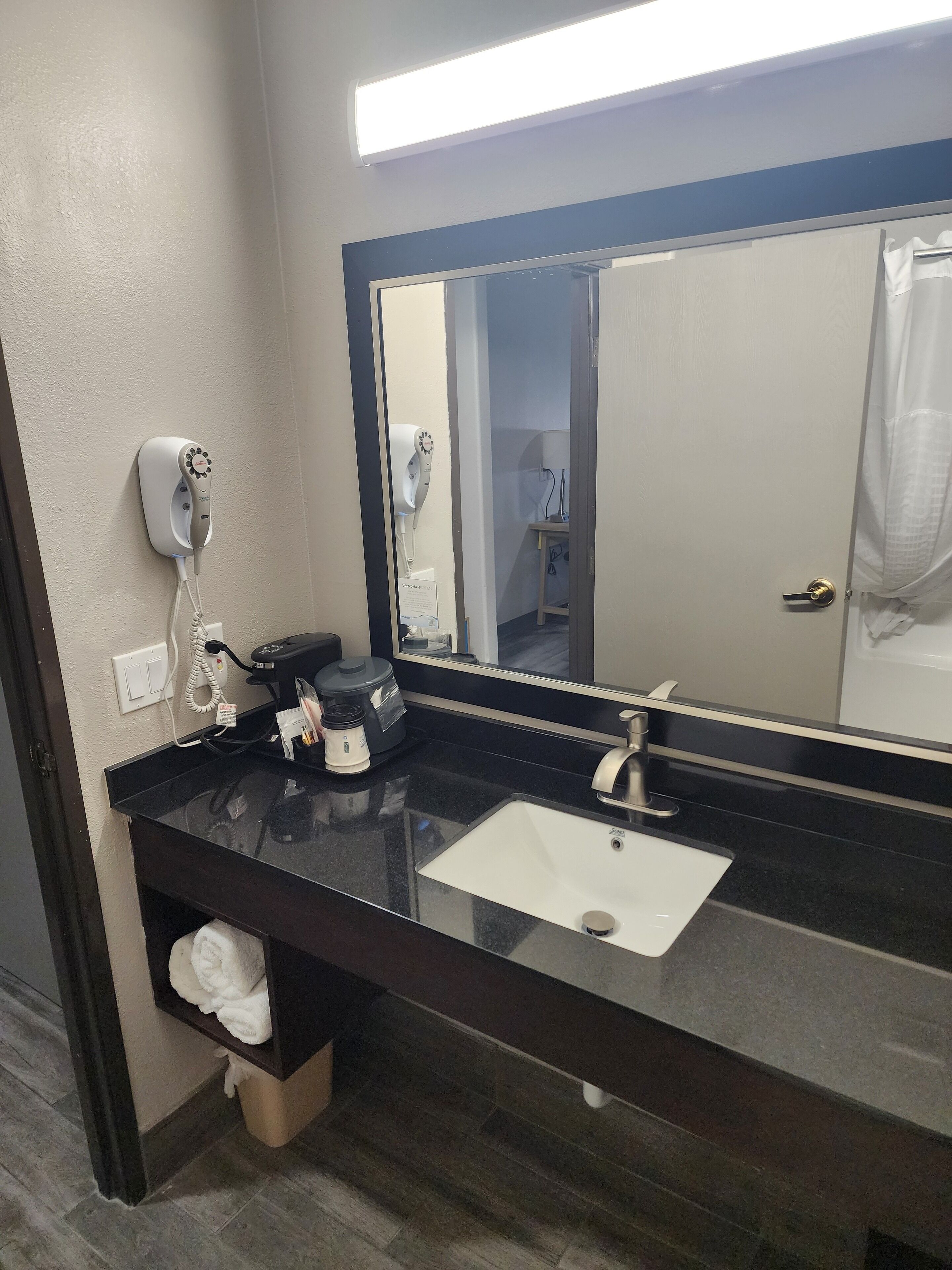 Casa de banho de hotel com bancada preta, lavatório branco, secador de cabelo e espelho.