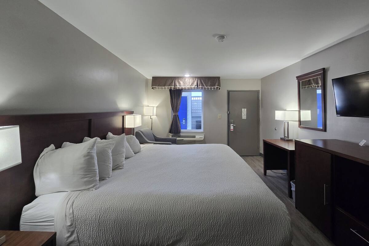 Quarto de hotel com uma cama grande, duas mesas de cabeceira com luminárias, uma televisão, uma escrivaninha e uma janela com cortinas.