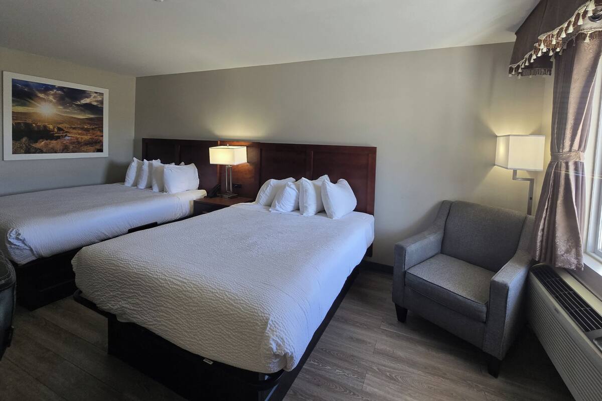 Quarto de hotel com duas camas, uma cadeira, mesa de cabeceira com abajur, quadro na parede e janela com cortinas.