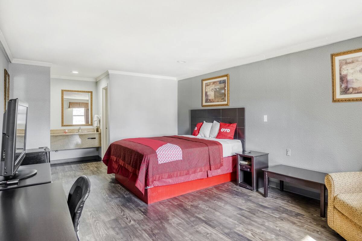 Quarto de hotel com cama, escrivaninha, cadeira, televisão e banheiro visível ao fundo.