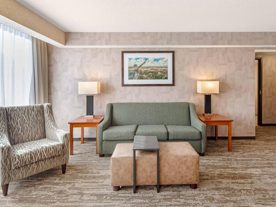 Quarto de hotel com poltrona estampada, sofá verde, um pequeno banquinho e duas luminárias de cabeceira, além de uma imagem emoldurada de uma vista urbana na parede.