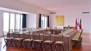Uma longa mesa de jantar posta para um evento formal, com cadeiras, bandeiras e uma grande janela.