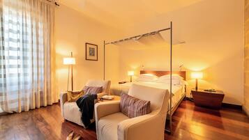 Quarto de hotel com cama de dossel, duas poltronas, piso de madeira e uma janela grande com cortinas.