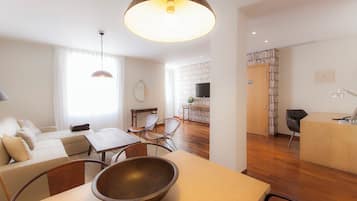 Uma sala de estar moderna com sofá, mesa de jantar e cadeiras. Há uma grande tigela de madeira sobre a mesa, um lustre pendente e uma estante com livros.