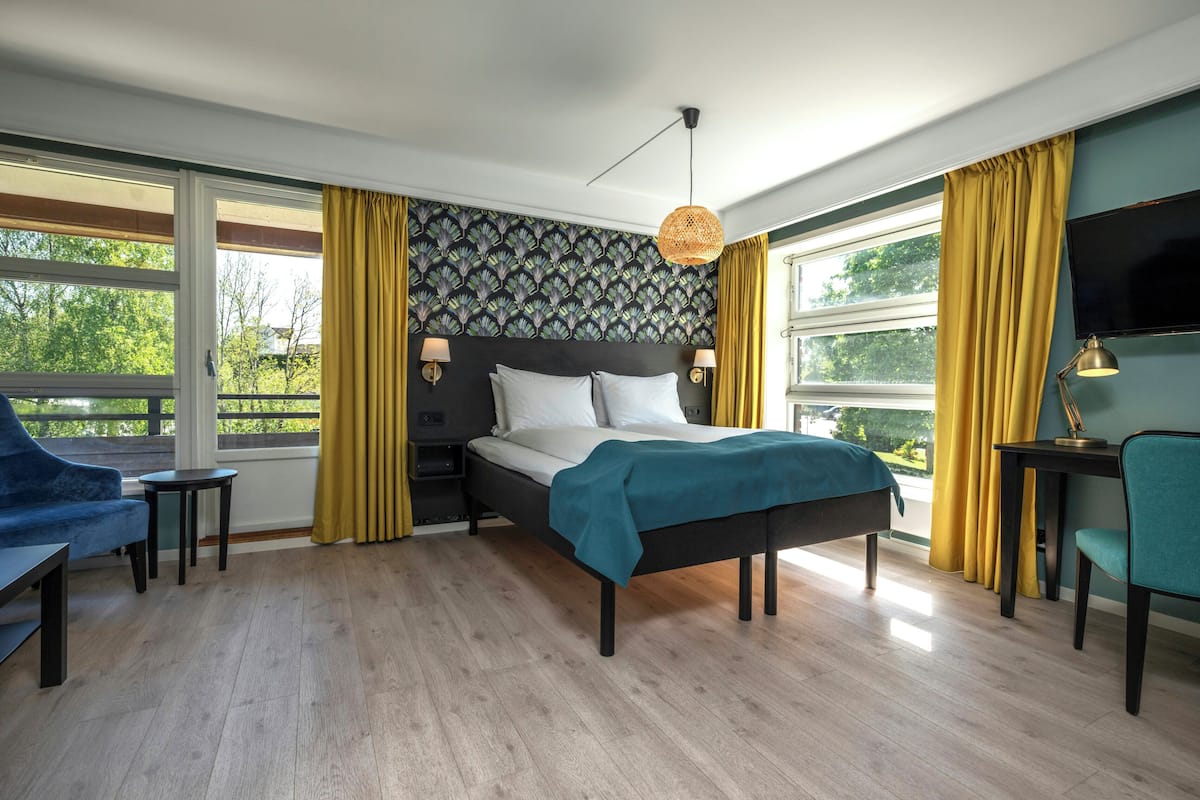 Quarto com cama, escrivaninha, cadeira, televisão e amplas janelas com cortinas.