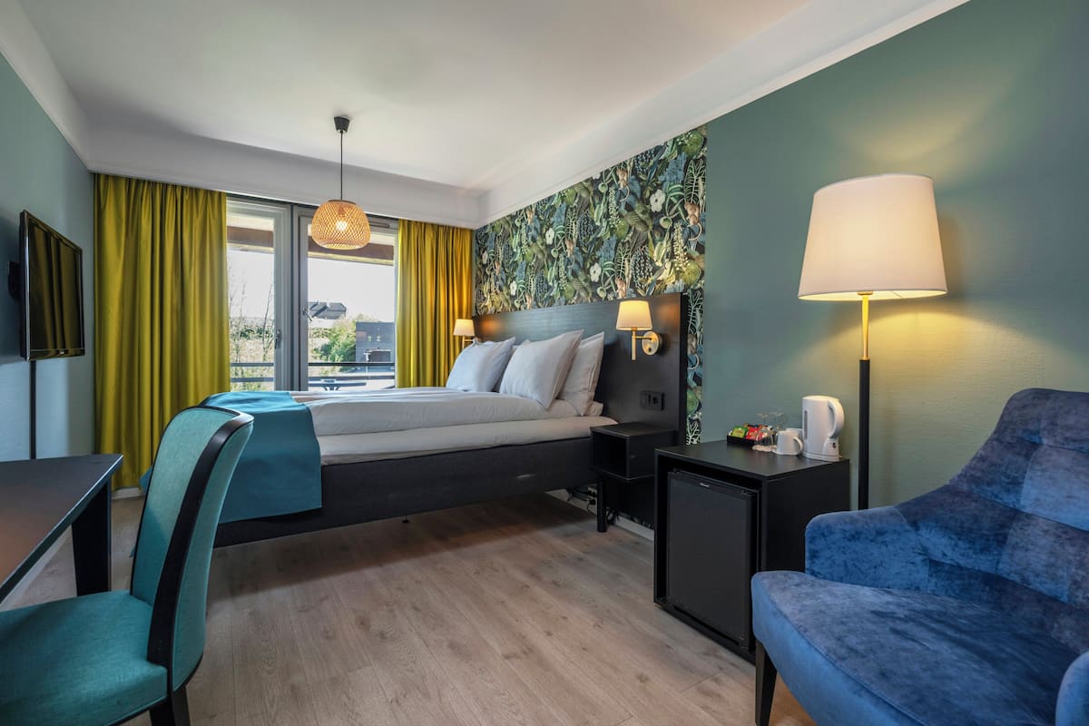 Quarto de hotel com cama, escrivaninha, cadeira, abajur e vista para o exterior.