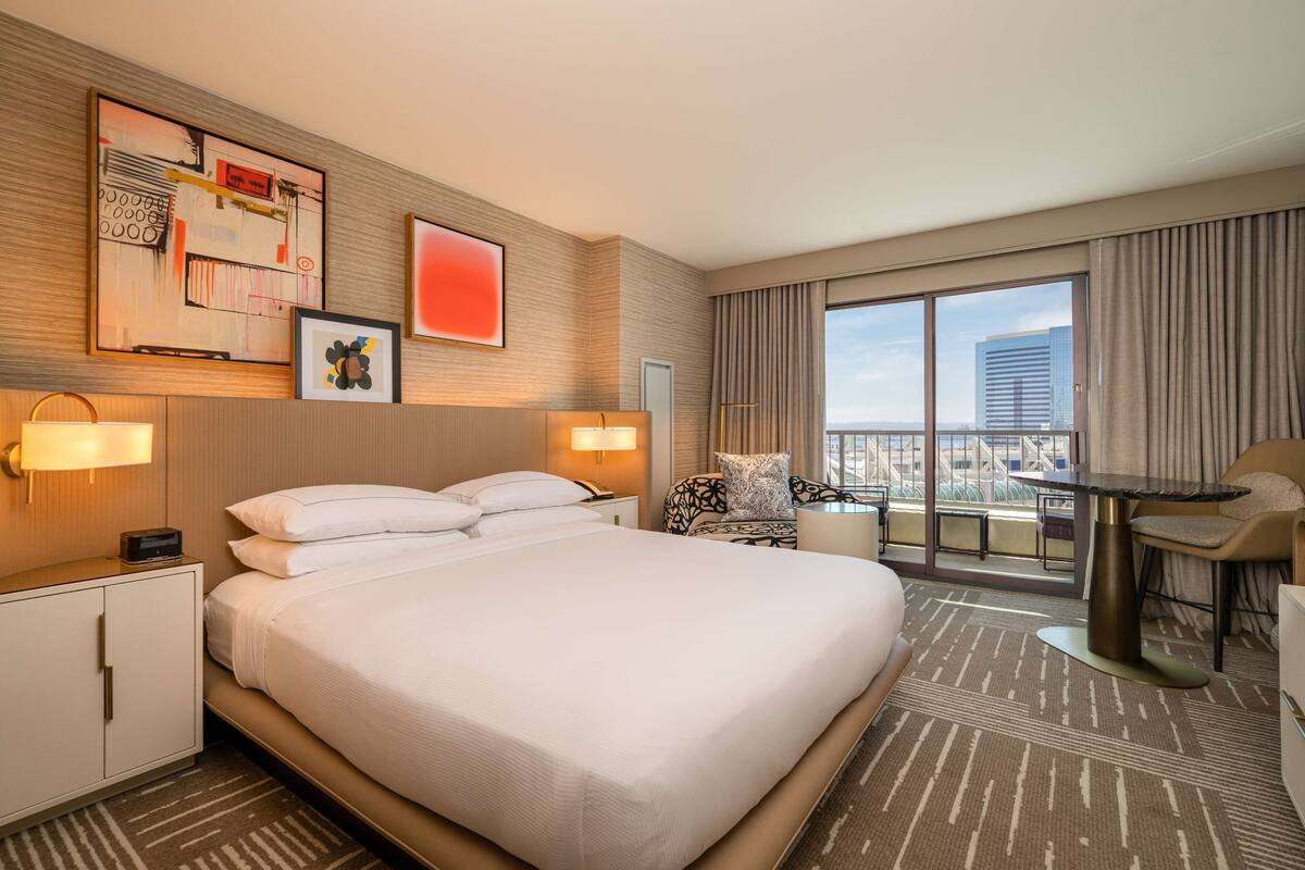 Quarto de hotel com uma cama grande, uma escrivaninha, uma cadeira, varanda com vista e obras de arte nas paredes.