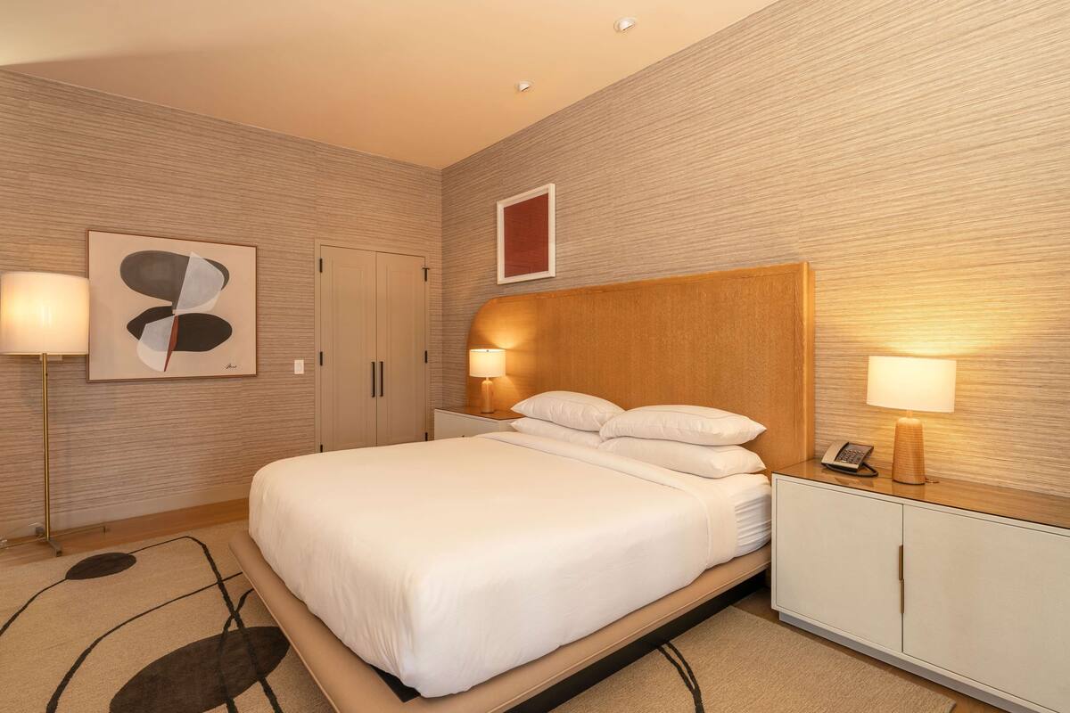 Quarto de hotel moderno com uma cama grande, duas luminárias de cabeceira, um painel de madeira na cabeceira e uma mesa de cabeceira pequena com um telefone, além de uma obra de arte abstrata emoldurada na parede.