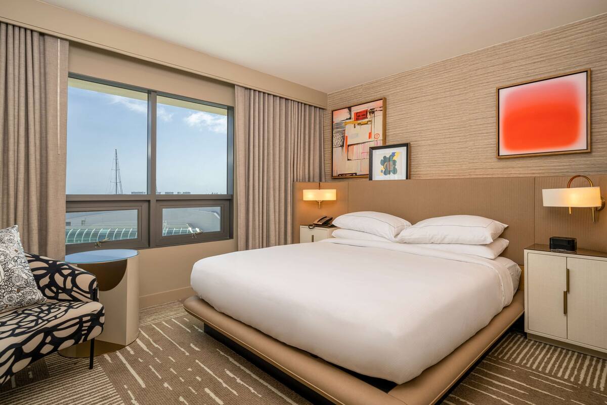 Quarto de hotel com uma cama grande, uma escrivaninha, uma cadeira, uma mesinha de cabeceira e vista para a cidade pela janela.