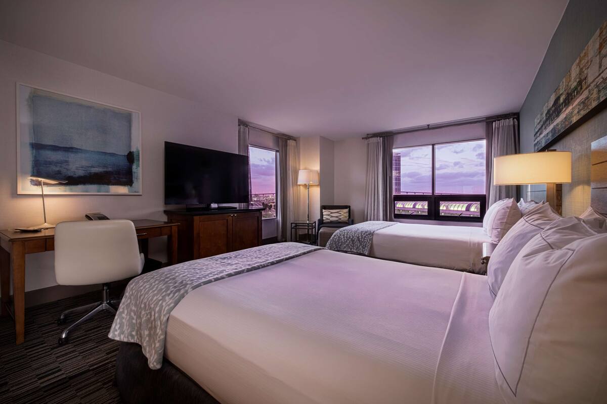 Quarto de hotel com duas camas, uma escrivaninha, uma cadeira, uma televisão e uma janela grande com vista.