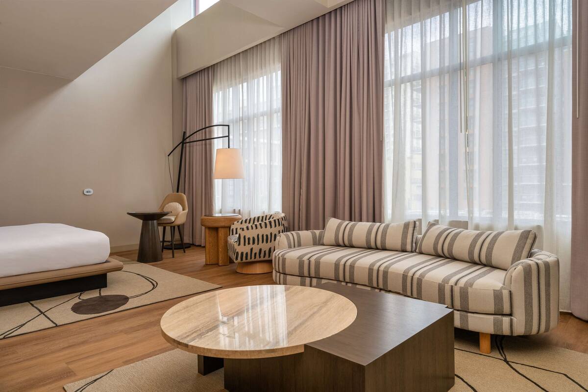 Quarto de hotel moderno com uma cama, um sofá, uma mesa de centro redonda e uma luminária.