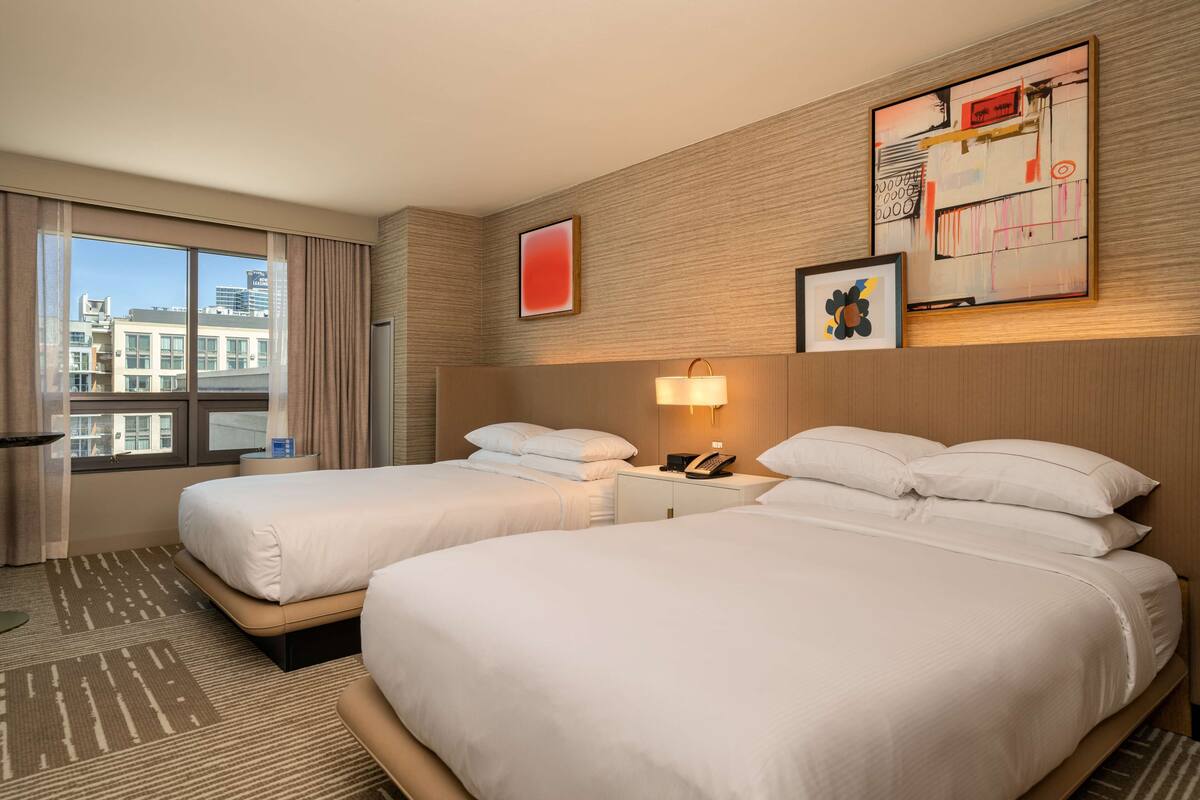 Quarto de hotel com duas camas, uma mesa de cabeceira com abajur, uma janela com cortinas e uma obra de arte na parede.