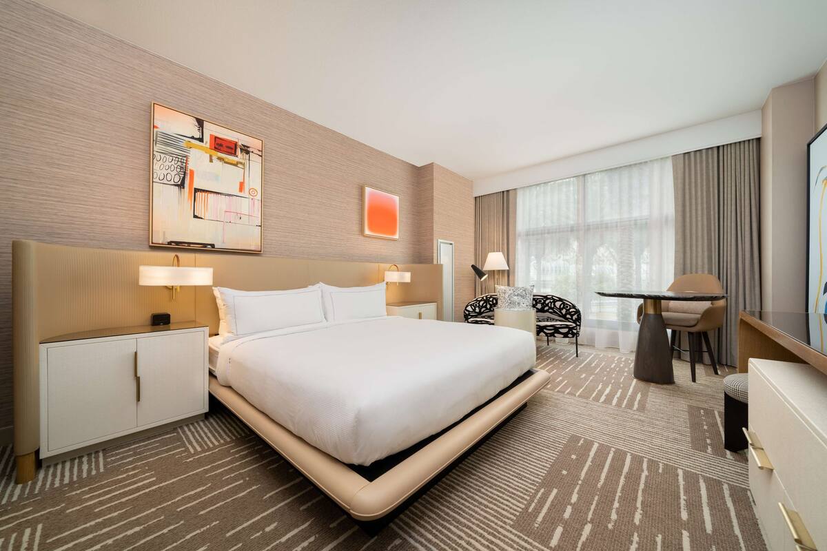 Quarto de hotel com uma cama grande, uma escrivaninha e uma cadeira. Há um quadro na parede e uma janela com cortinas.