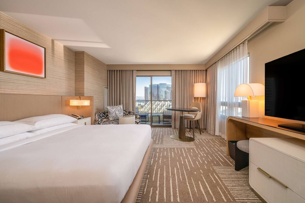 Quarto de hotel com cama grande, escrivaninha, televisão e varanda com vista para a cidade.