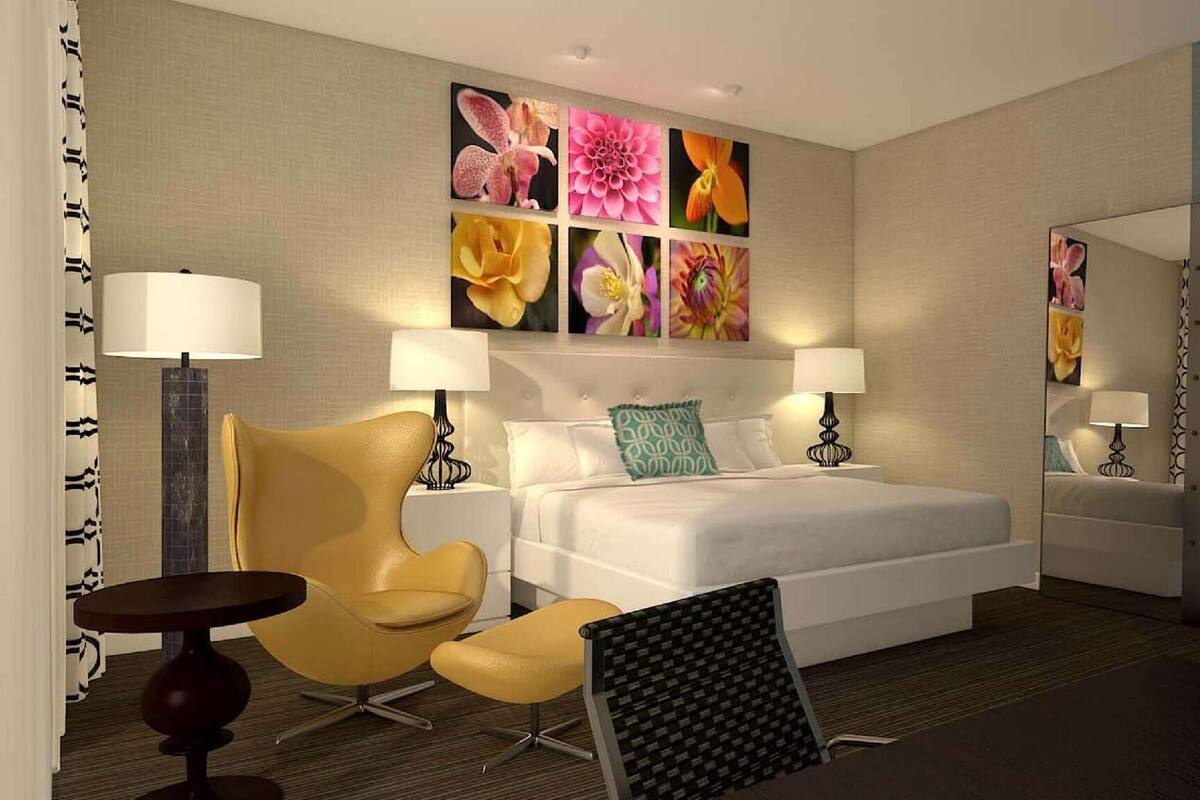 Quarto de hotel com uma cama, duas luminárias de cabeceira, uma cadeira, uma mesa e uma grande obra de arte floral na parede.