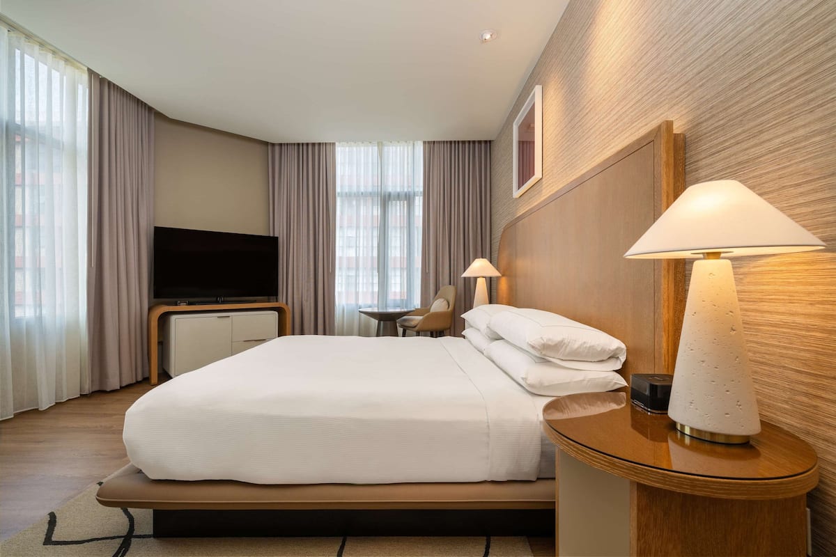 Quarto de hotel com uma cama grande, televisão, mesa de cabeceira com abajur e uma pequena mesa com cadeira.