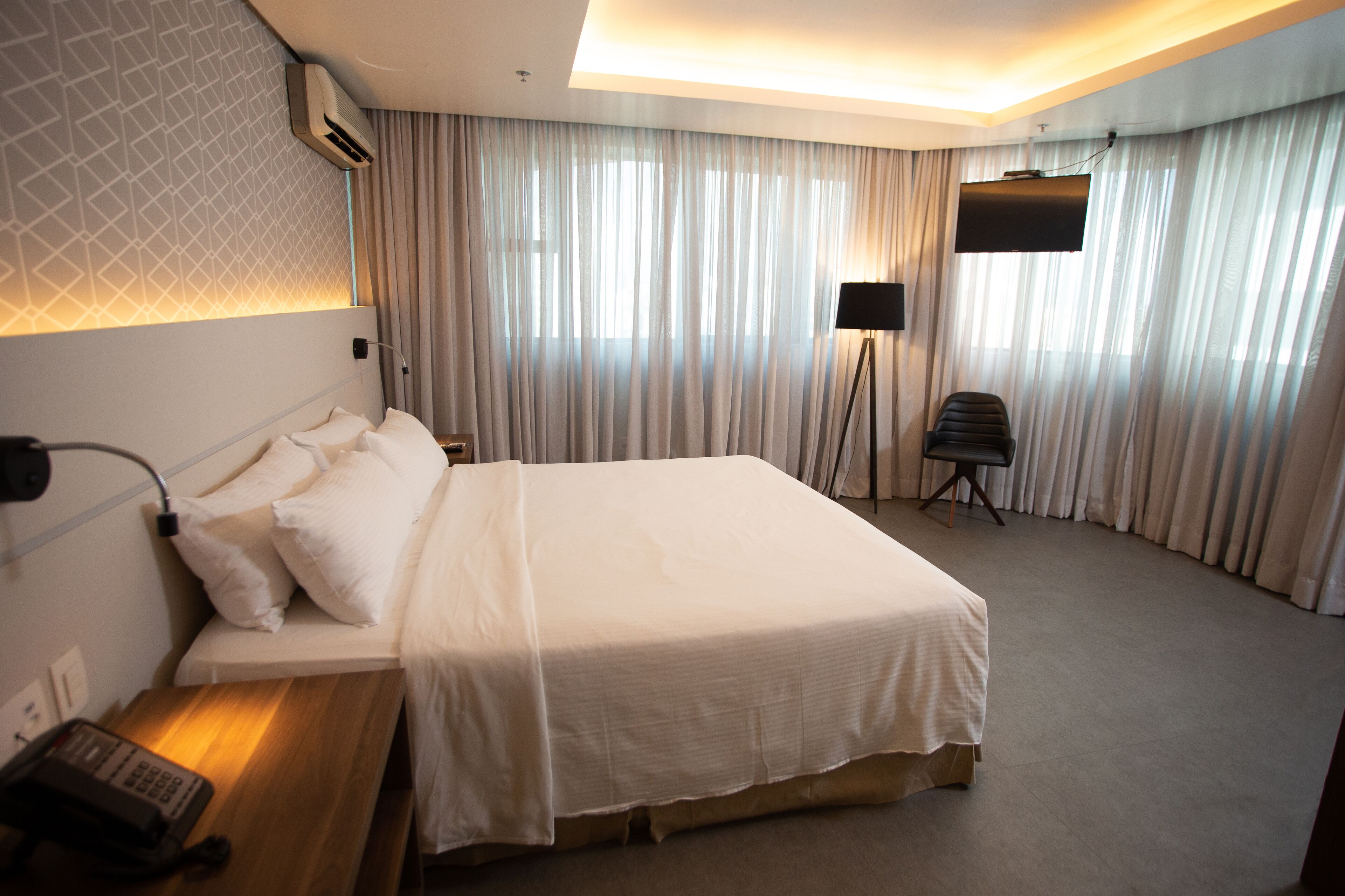 Quarto de hotel com uma cama grande, mesa de cabeceira com telefone, abajur e televisão na parede.