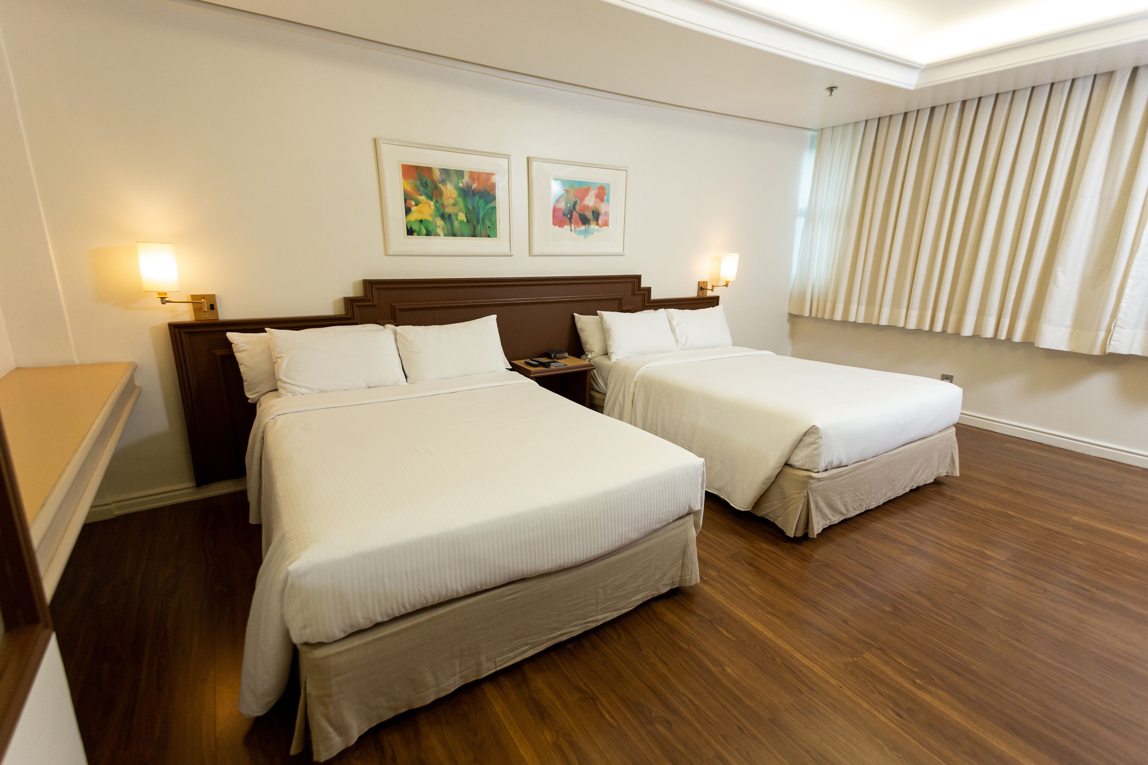 Quarto de hotel com duas camas, soalho de madeira, mesa de cabeceira, candeeiro e duas obras de arte emolduradas na parede.