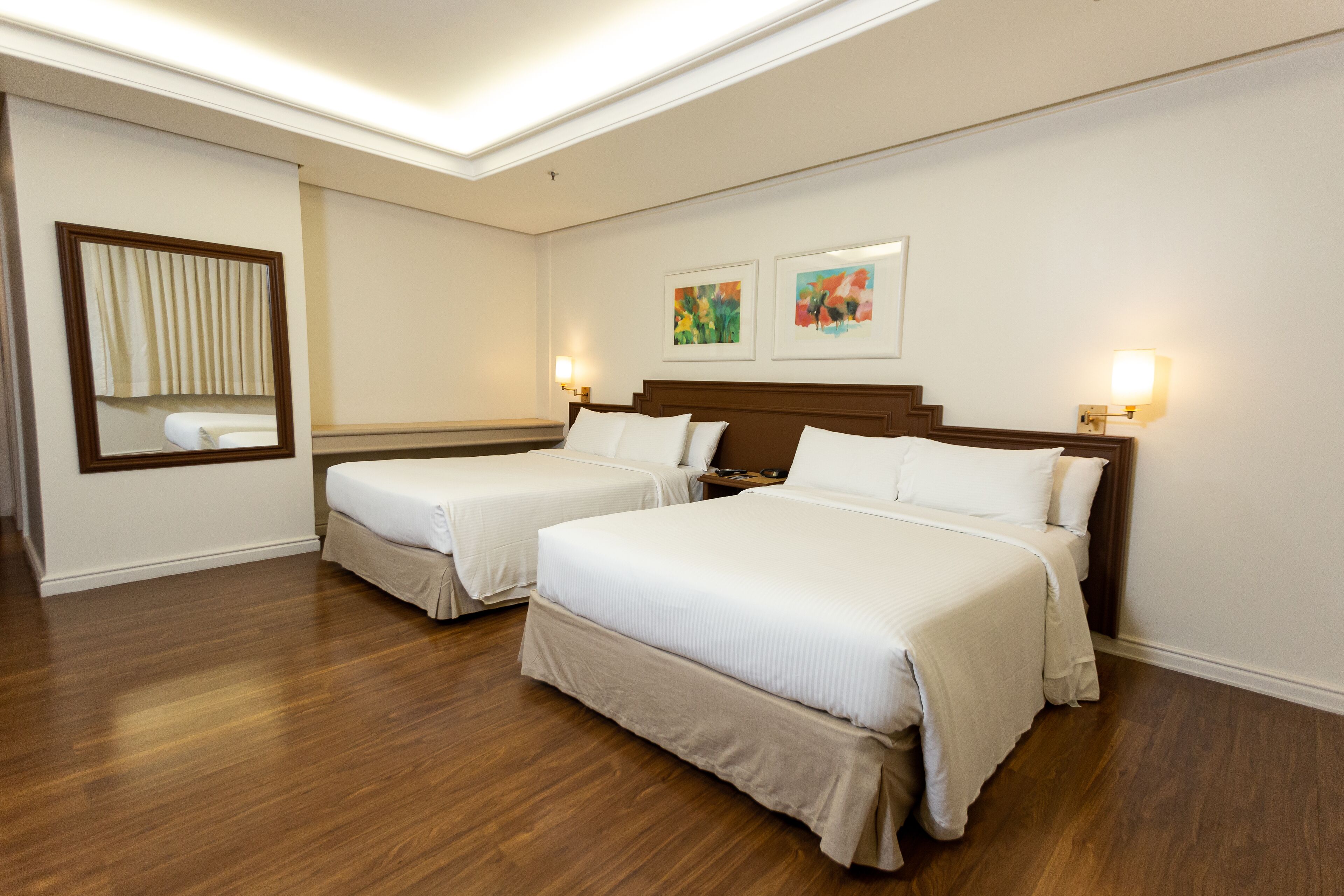 Quarto de hotel com duas camas, piso de madeira, um espelho grande e duas luminárias fixadas na parede.