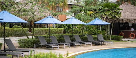 Uma área à beira da piscina com espreguiçadeiras e guarda-sóis, cercada por vegetação e um edifício com telhado de palha.