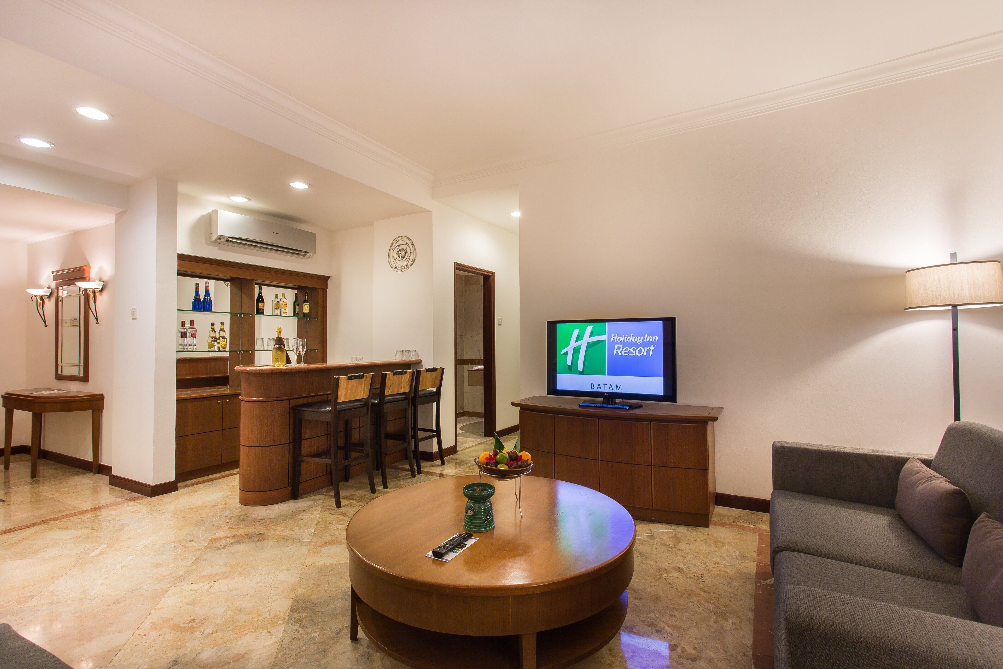 Um hall de hotel com uma mesa redonda de madeira, um sofá, uma televisão exibindo um resort, um bar e uma área de refeições.