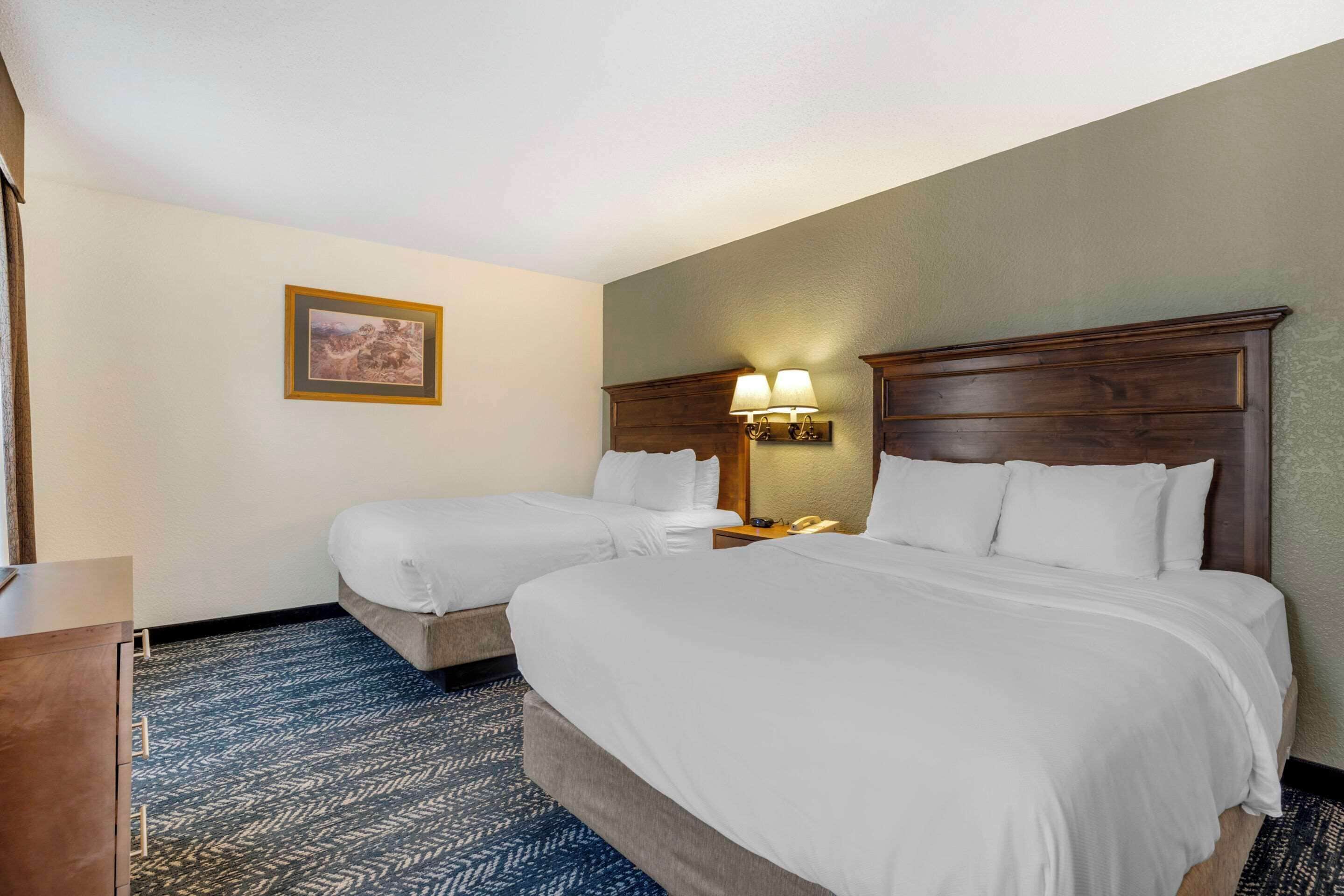Quarto de hotel com duas camas, uma mesinha de cabeceira com abajur, um quadro na parede e um tapete estampado.